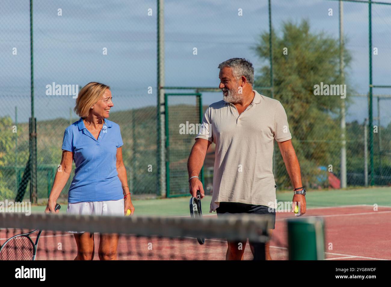 closeup elderly couple smiles and chats as they walk towards the tennis court, the net in the ...