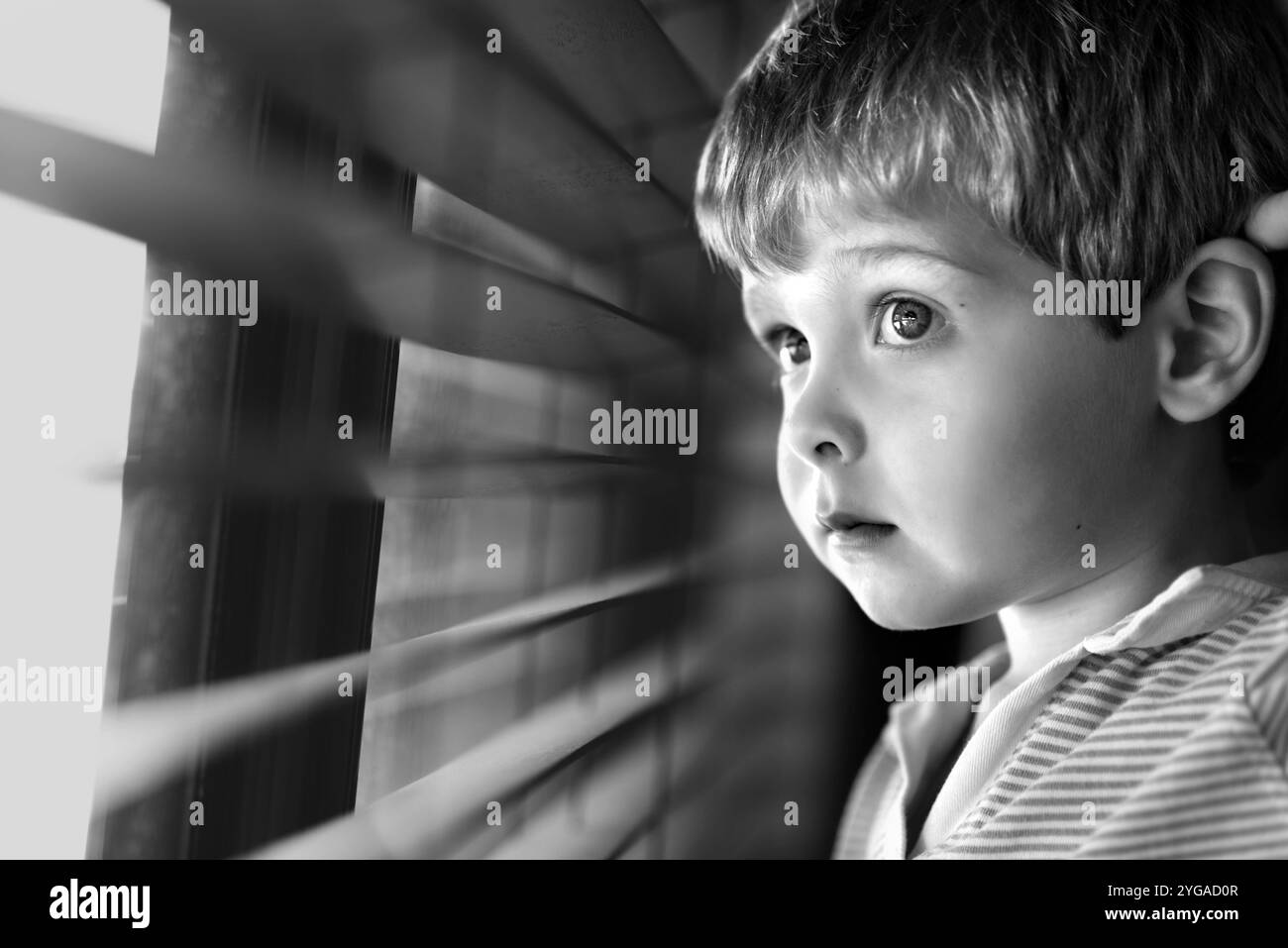 Young boy gazing out through window blinds in black and white, deep in ...