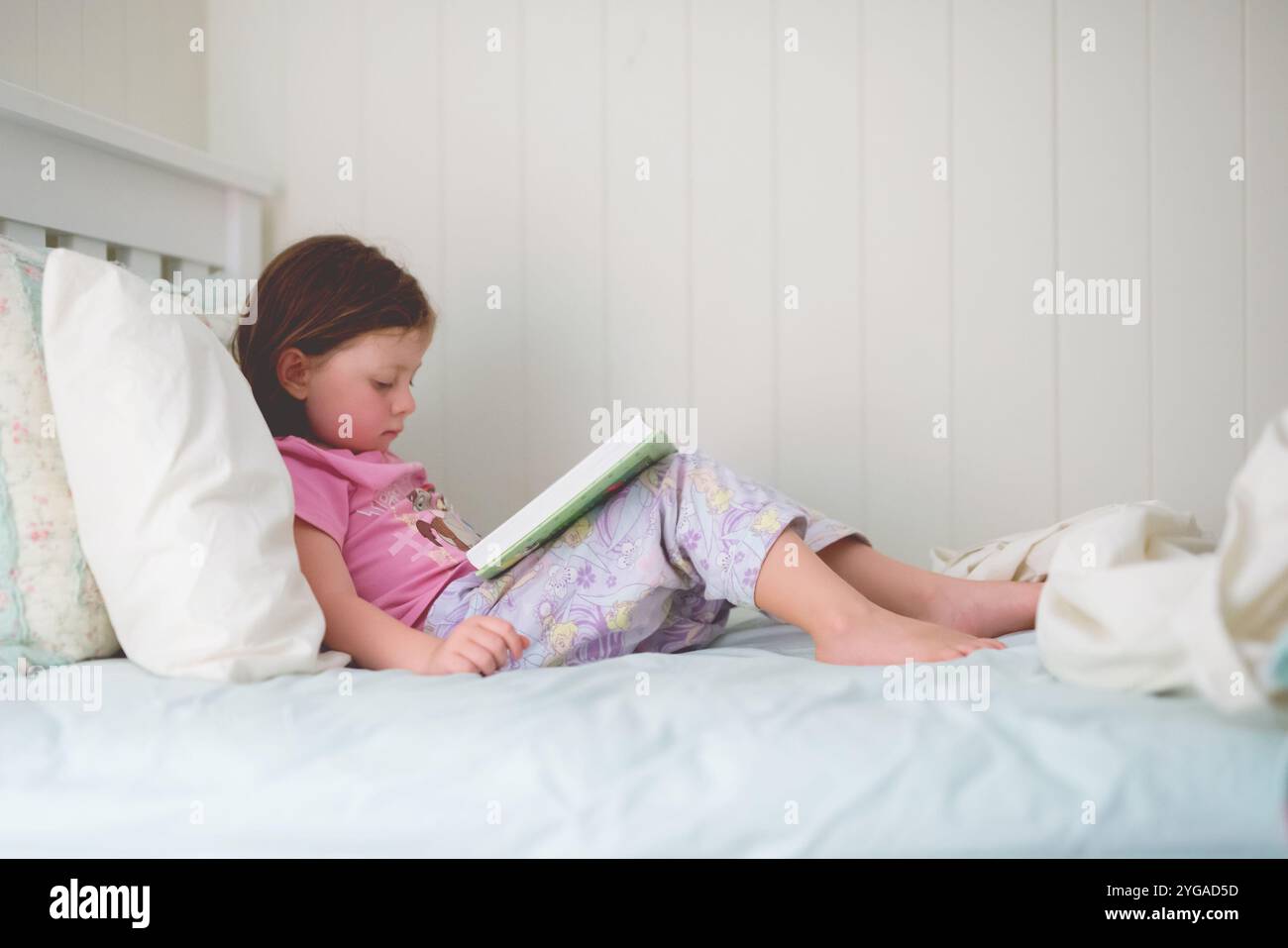 Young girl sitting on her bed in pyjamas reading a book in a cosy ...