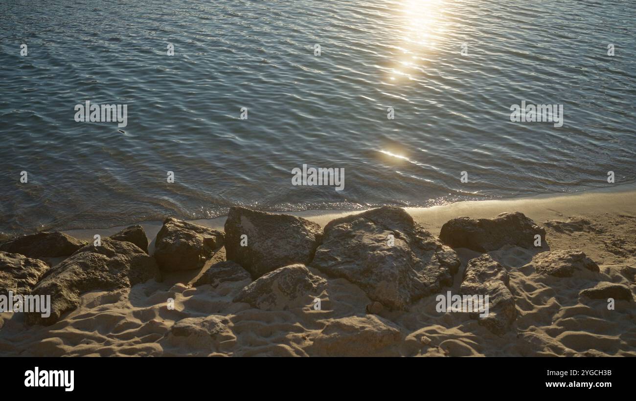 Serene shoreline with rocks and sandy beach as the sun sets reflecting ...