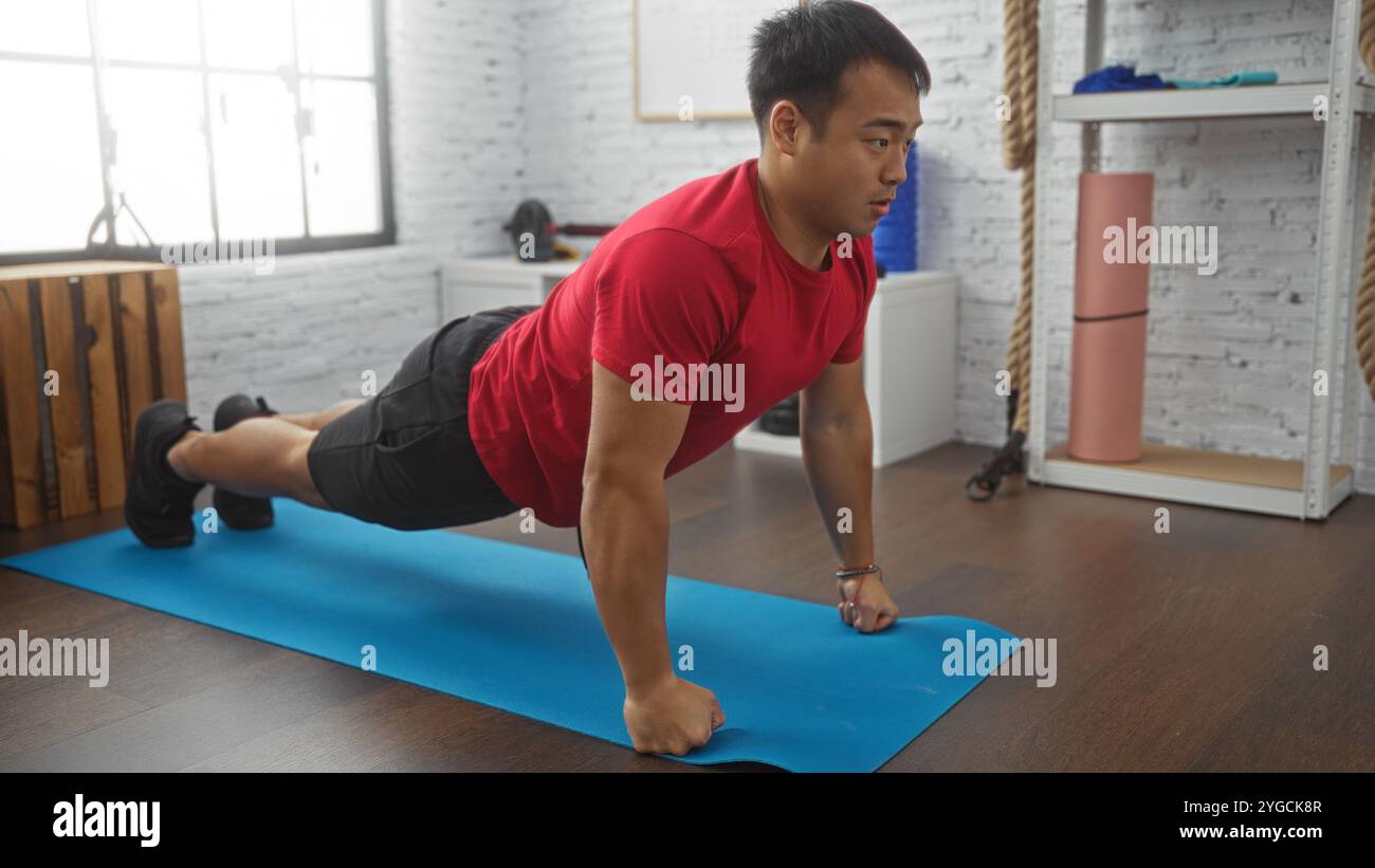 A young chinese man performing push-ups on a blue mat in a modern gym, showcasing his fitness in ...