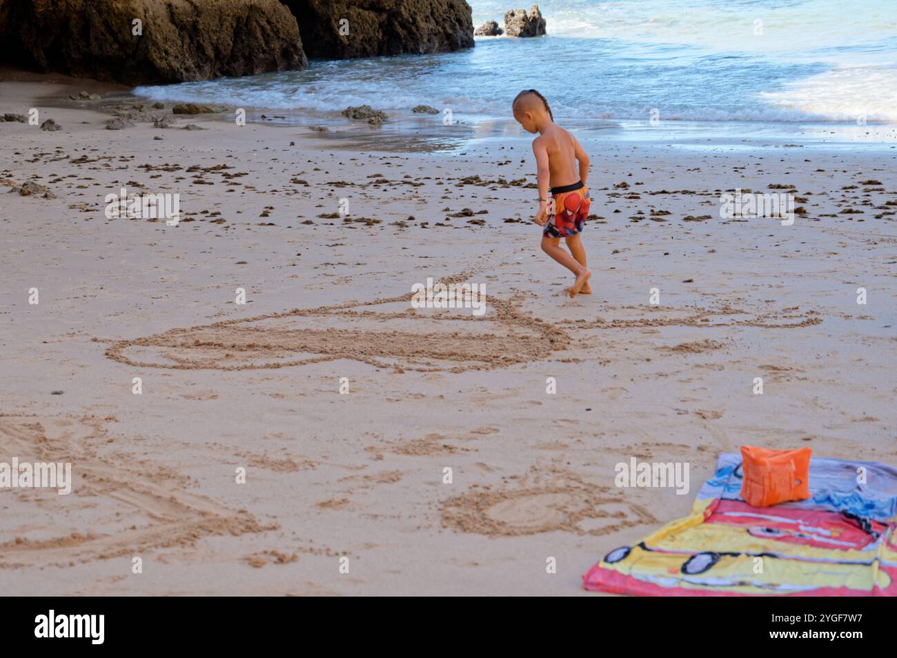 A young boy drawing shapes in the sand on the sandy shores of Praia da ...