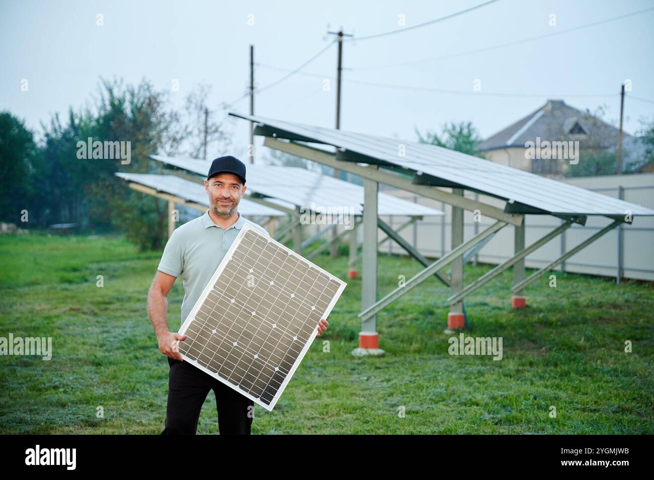 Man holds solar panel while standing in solar farm. Smiling guy wearing ...