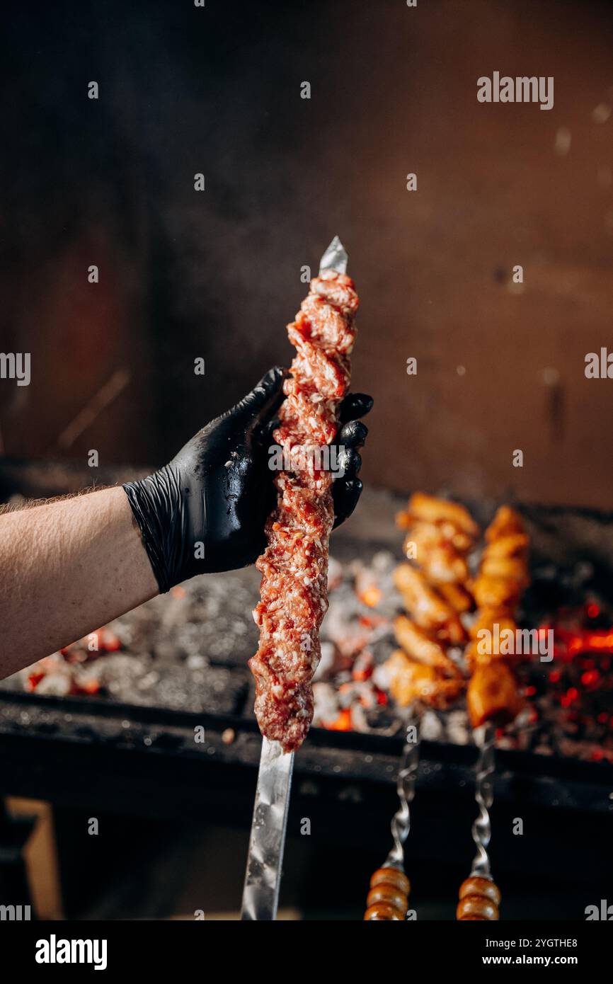 Chef preparing minced meat kebab skewer for grilling over open flame, a ...