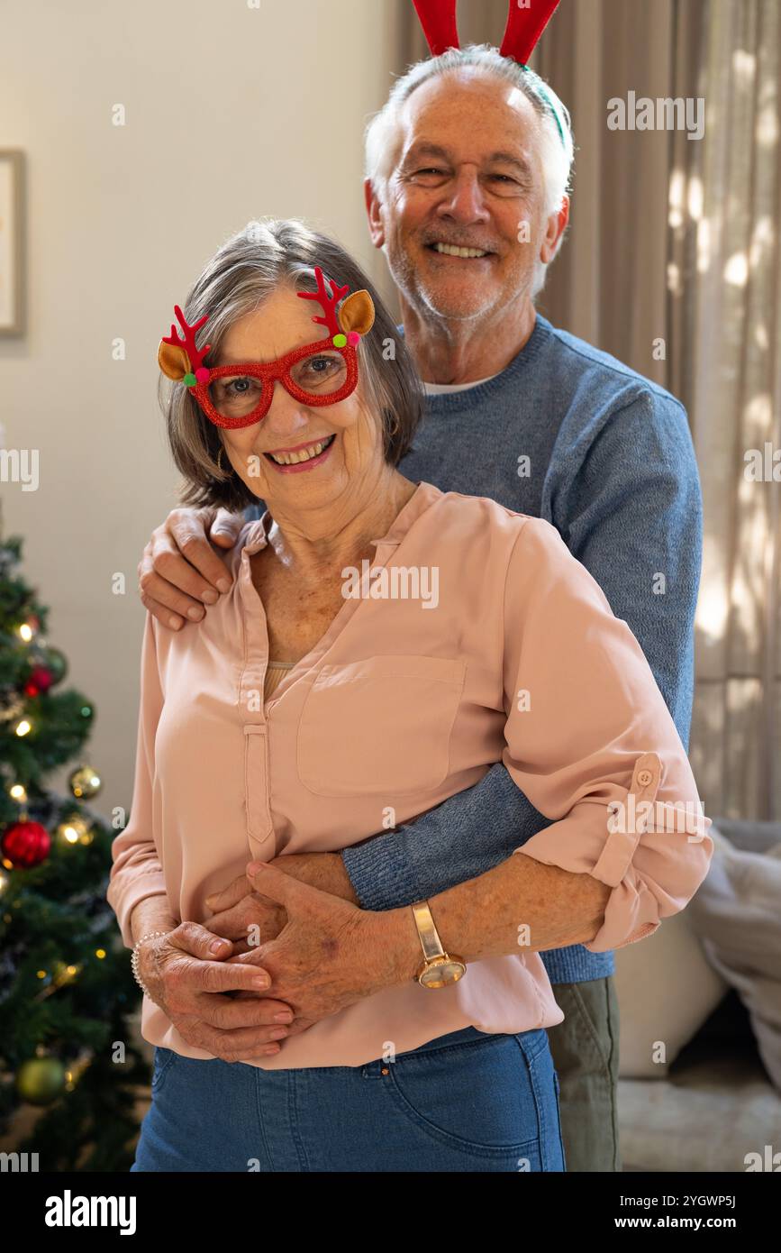 Senior couple wearing festive accessories smiling by Christmas tree at ...