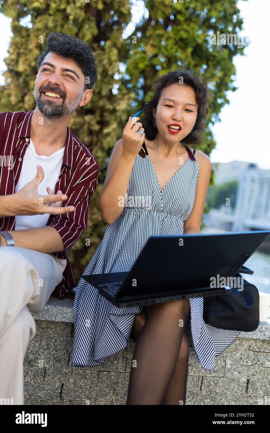 Two colleagues relaxing outside with a laptop, laughing and enjoying a ...