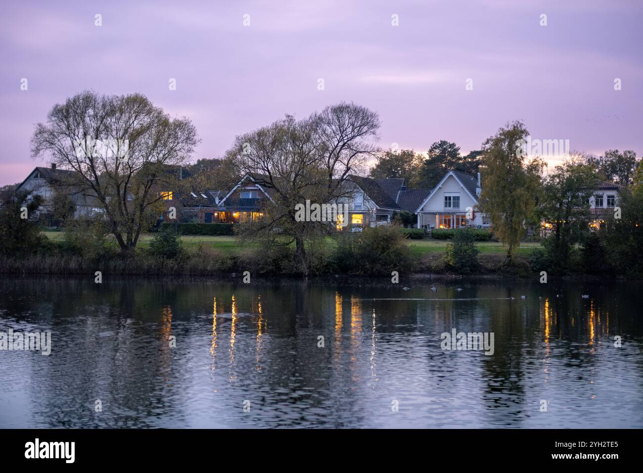 Serene lakeside view of cozy houses with illuminated windows at dusk, reflecting on calm water ...