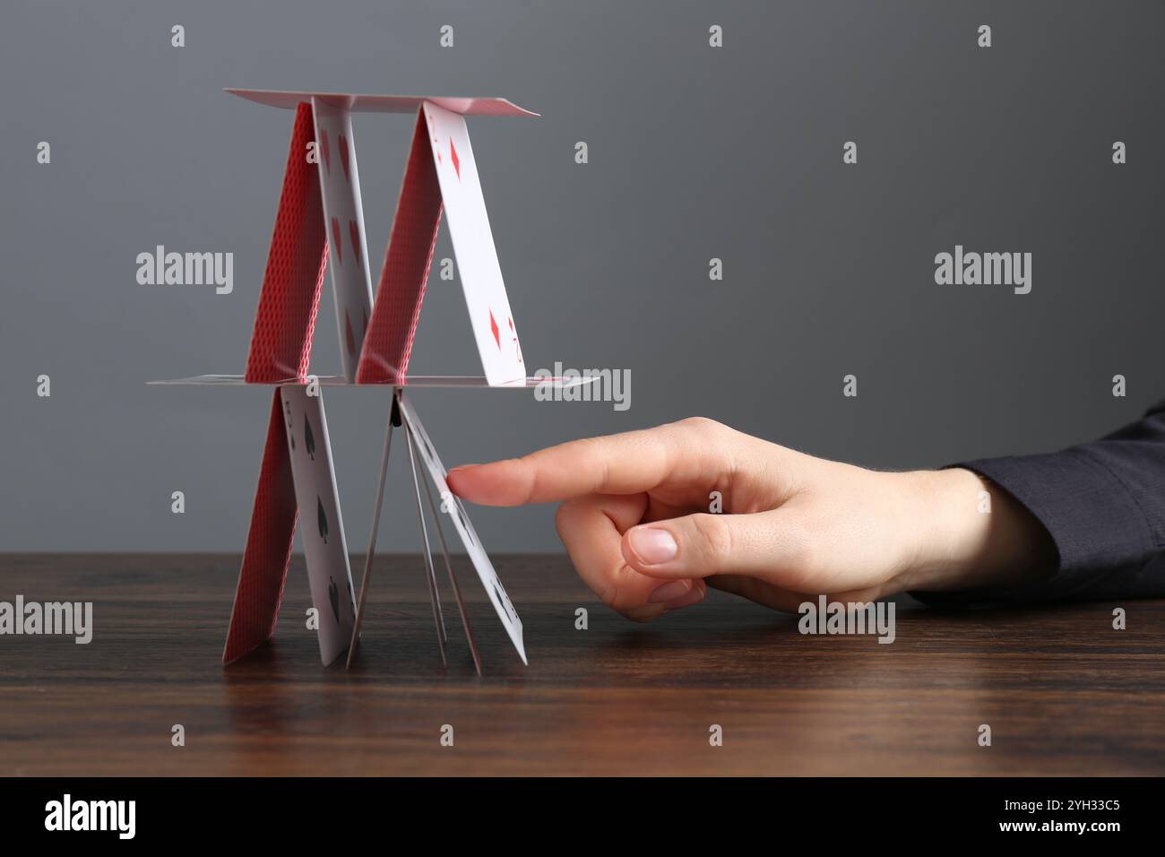 Woman destroying house of playing cards at wooden table against grey ...