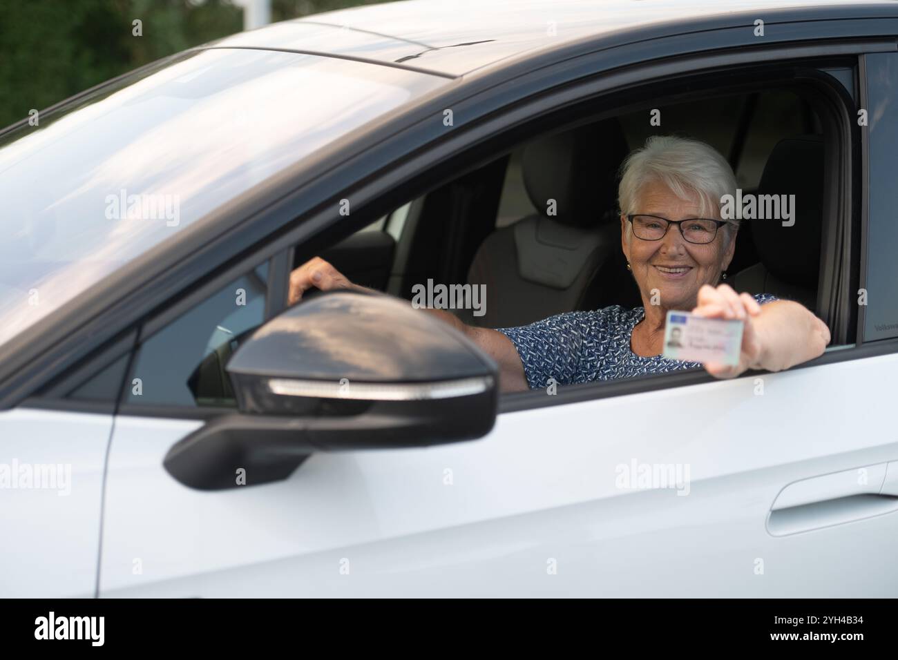 A happy elderly woman in the driver’s seat of a white car holds her driver’s license out the ...