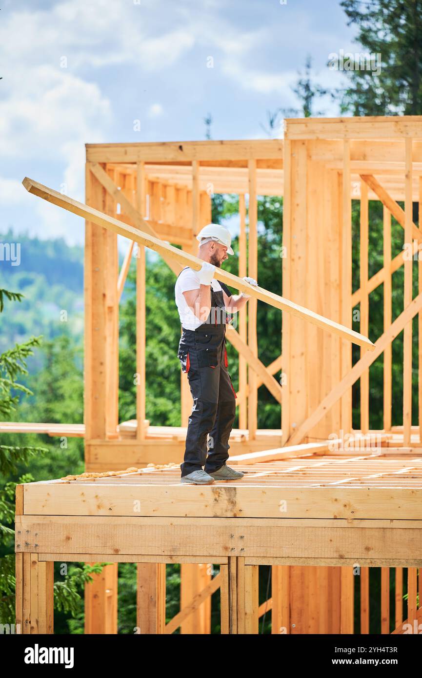Carpenter builds wooden frame house near woods. Bearded man holds large ...