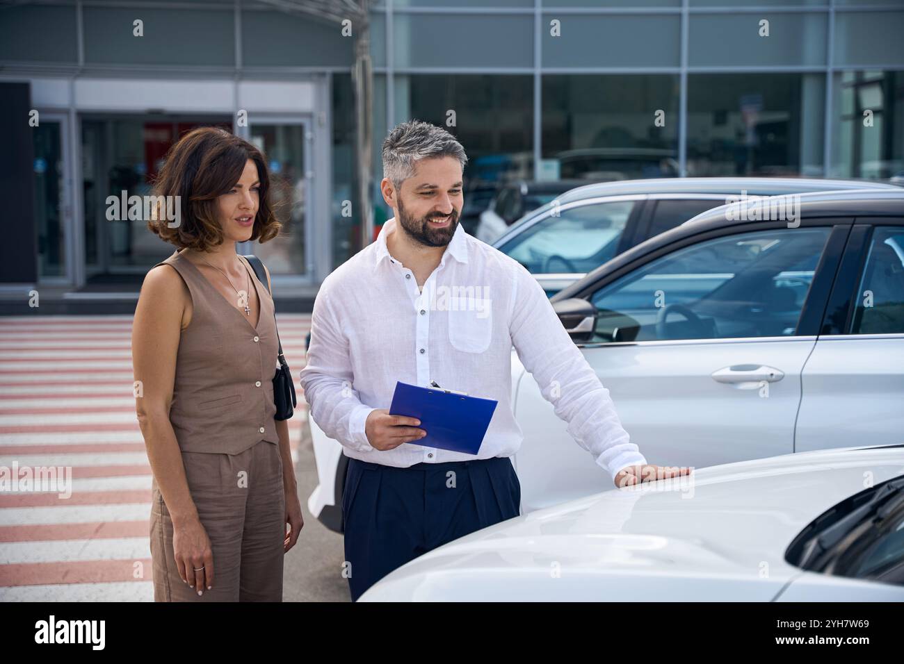 Salesman manager with file folder standing near new car with female ...