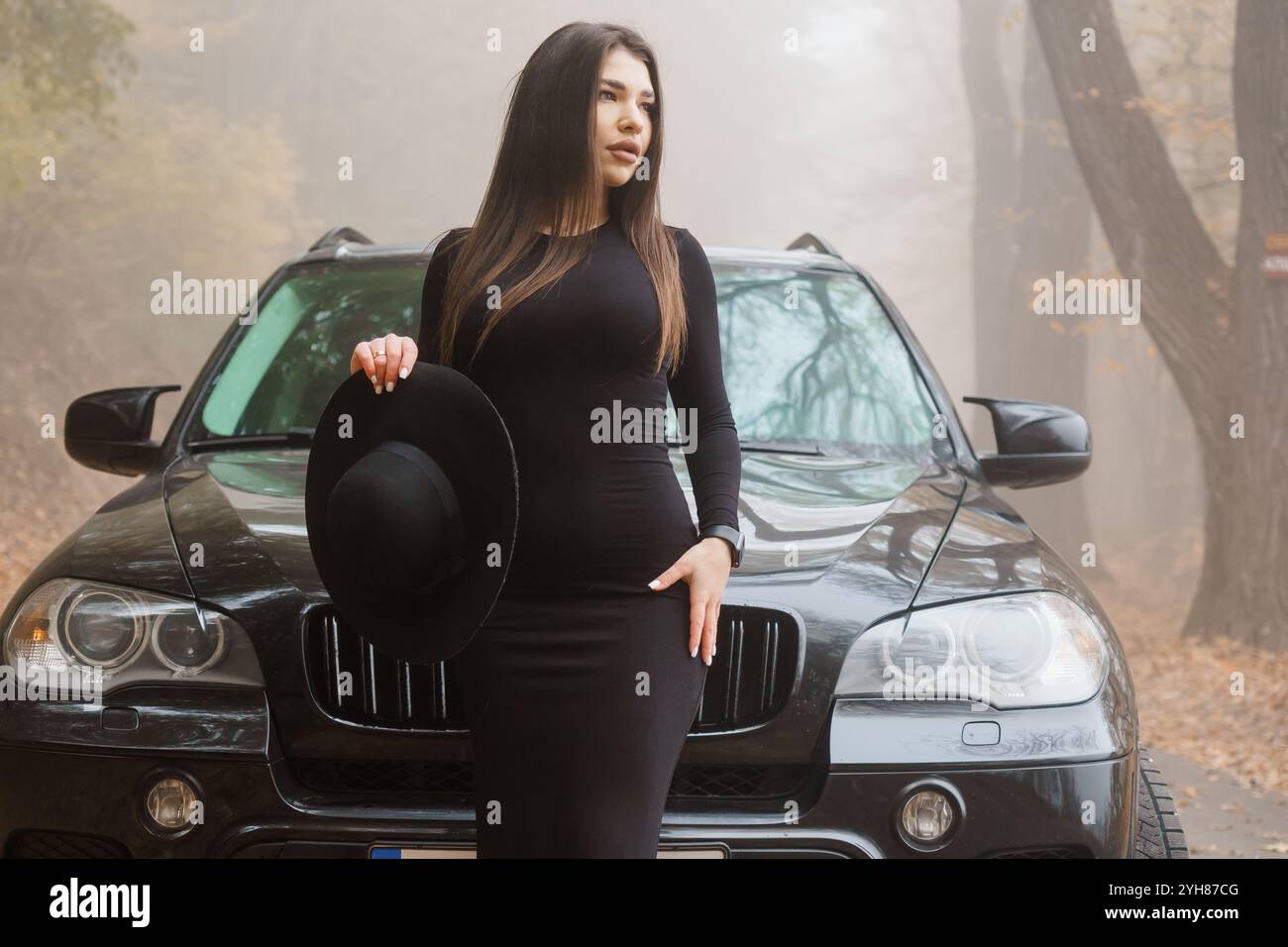 Elegant Woman in Black Dress Posing with Hat in Front of Luxury SUV in ...