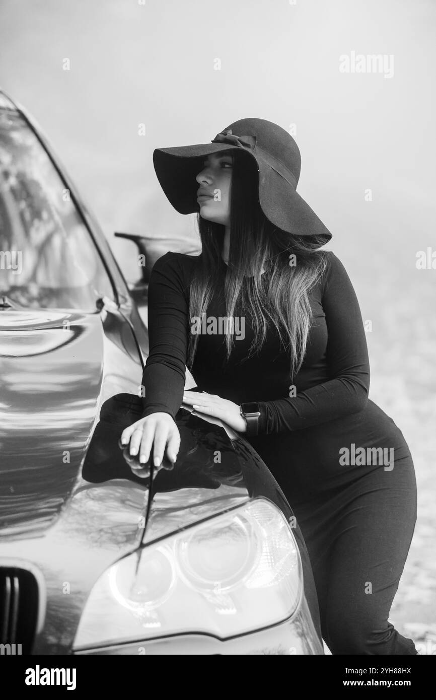 Elegant Woman in Black Dress and Wide-Brim Hat Posing by Luxury Car in ...