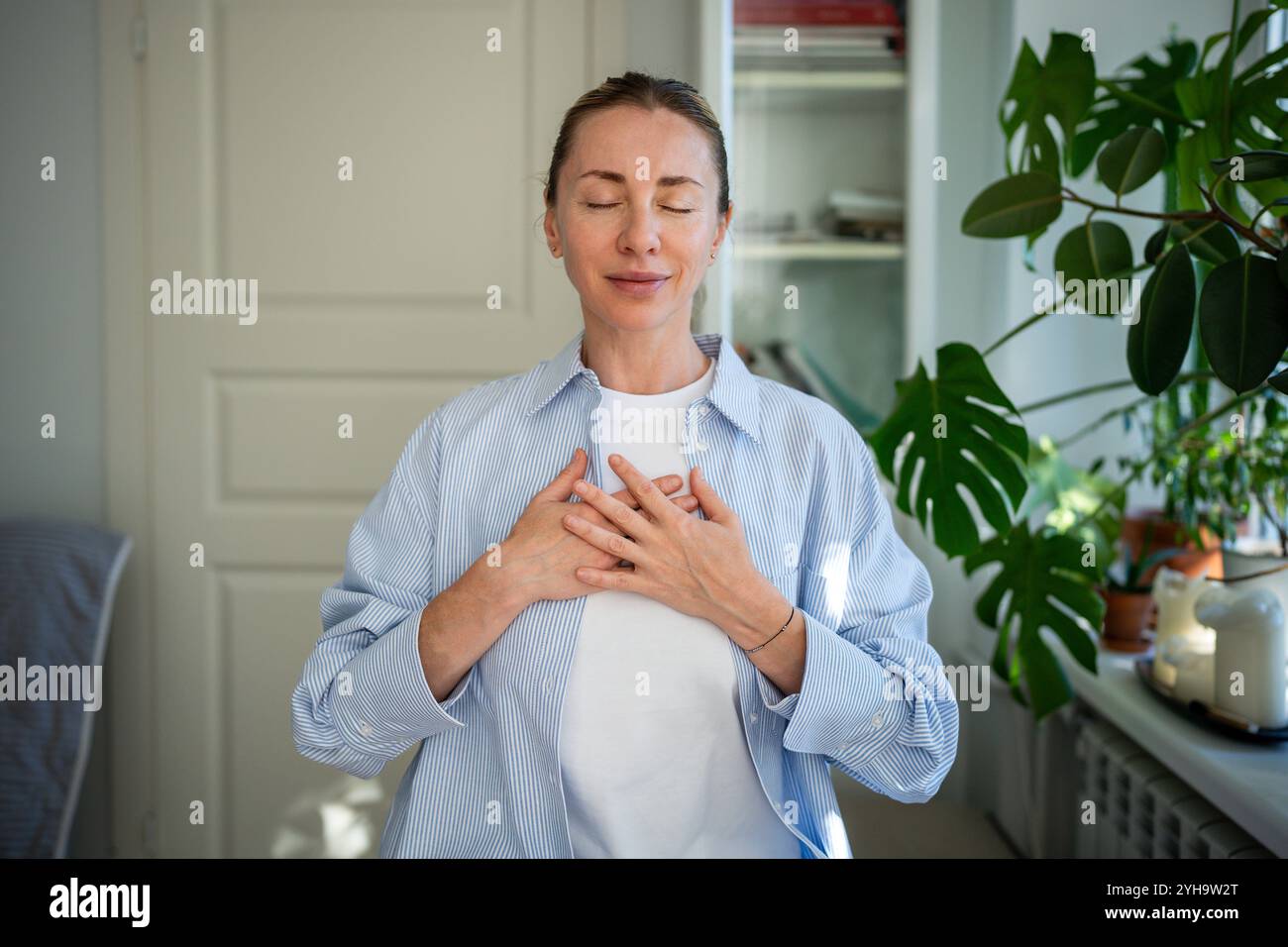 Peaceful woman standing with closed eyes, folding one's arms on chest doing breathing exercises ...