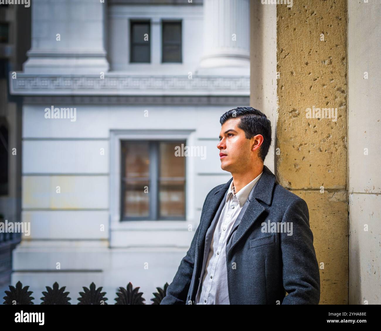 A man in a dark coat leans against a textured stone wall, gazing ...