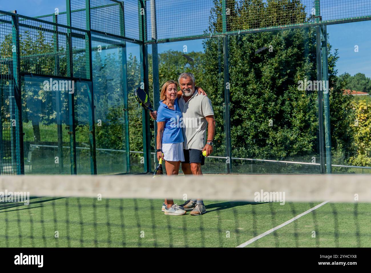 senior couple embraces on the padel court, looking at the camera with ...