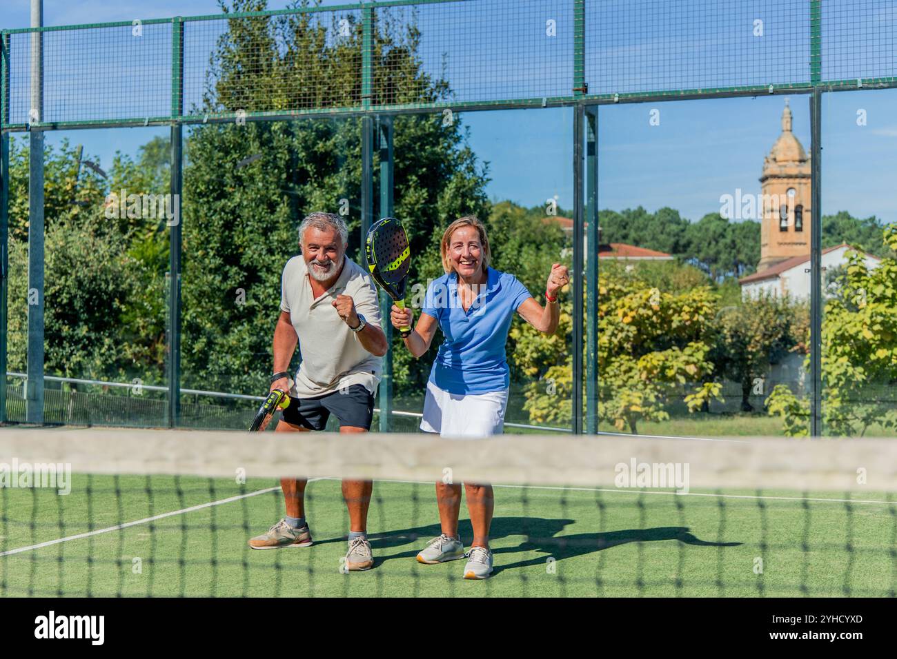 joyful elderly padel couple stands on the court, raising their fists ...