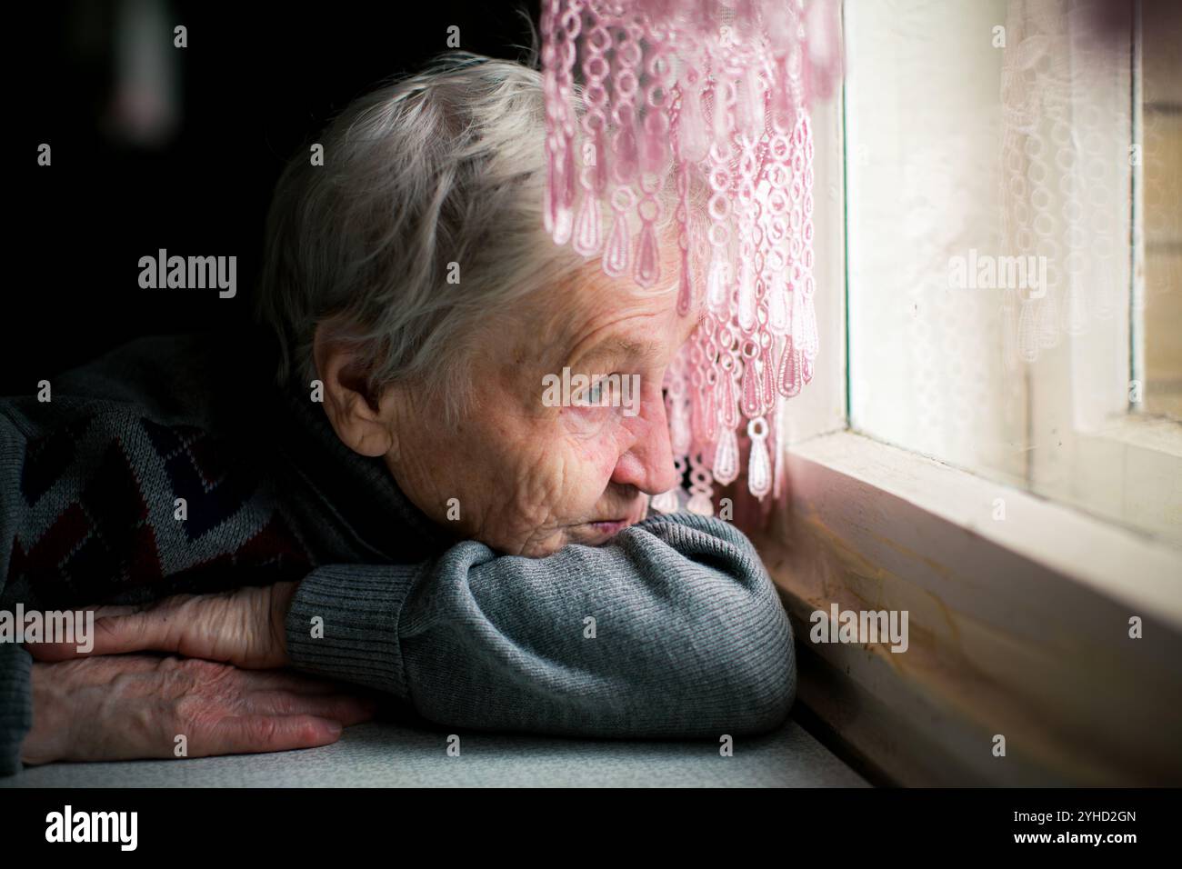 An elderly woman looks sadly out the window of her home, her eyes ...