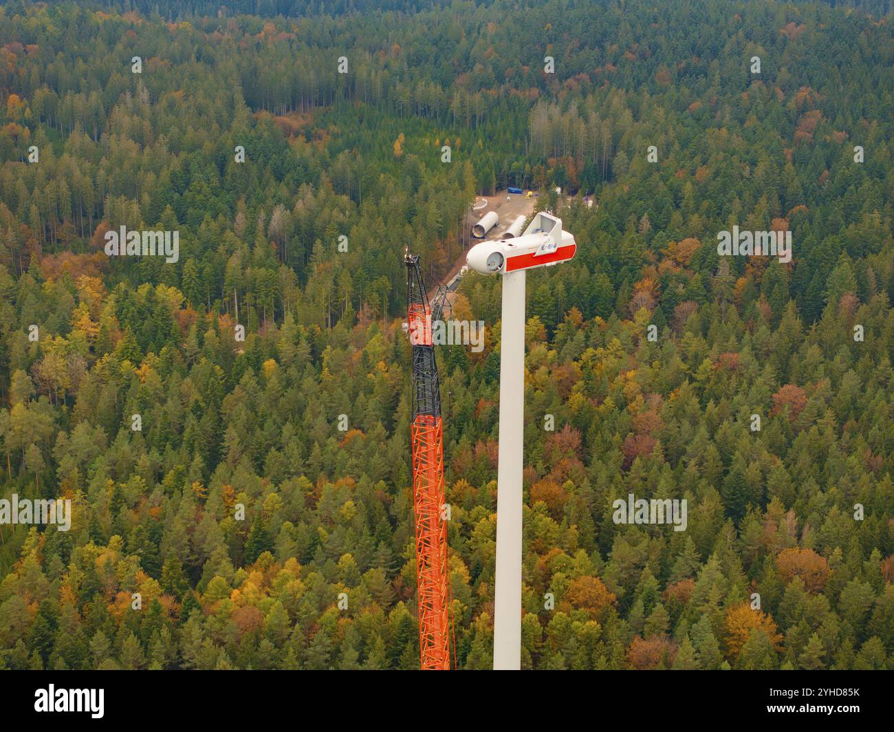 A crane assembles the upper part of a wind turbine above an autumnal ...