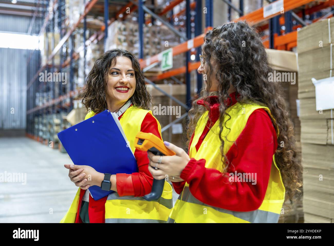 Two young women talking while taking inventory in a distribution warehouse using clipboard and ...