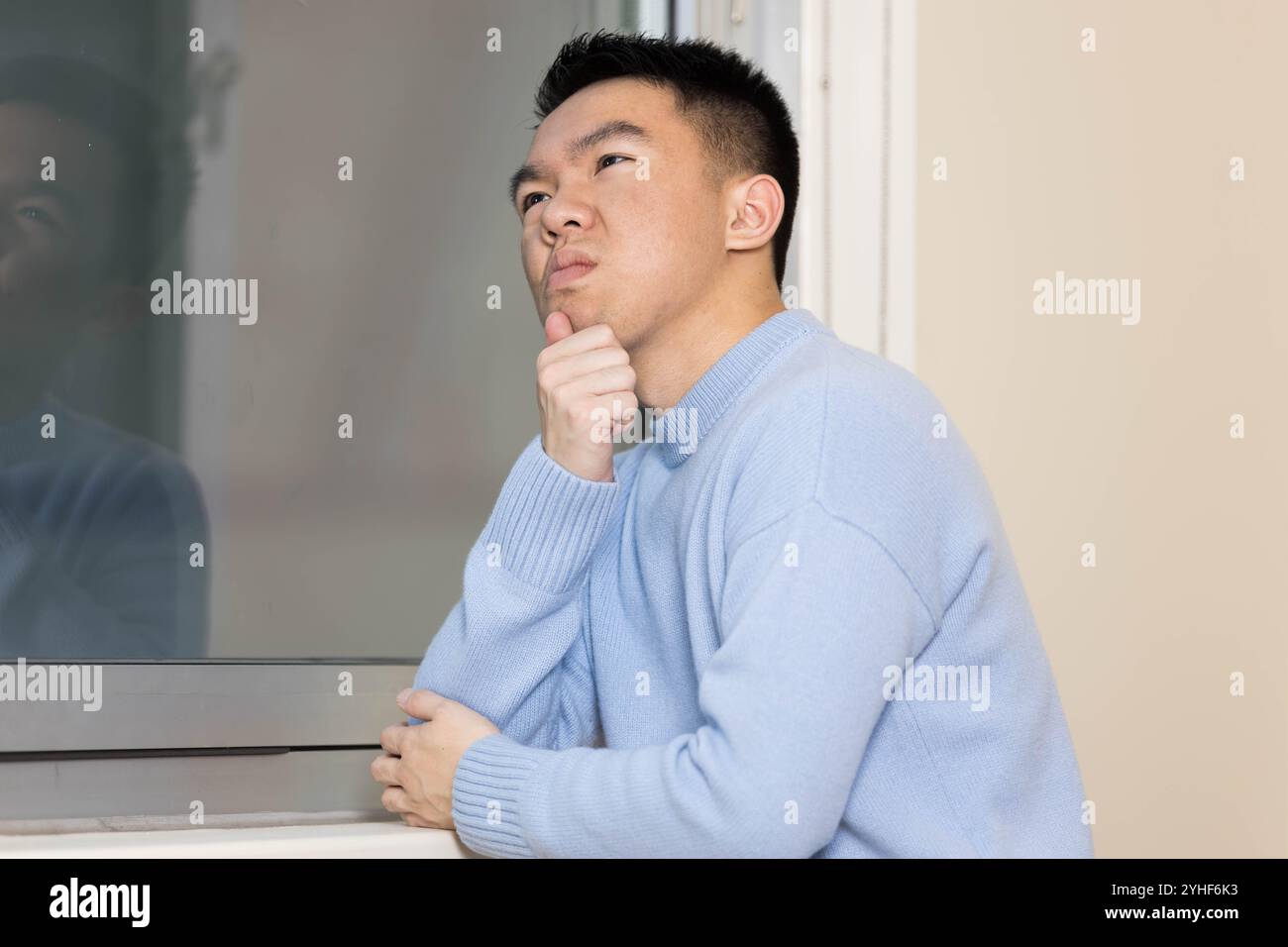 A young man is contemplating thoughtfully in front of a window, hand on ...