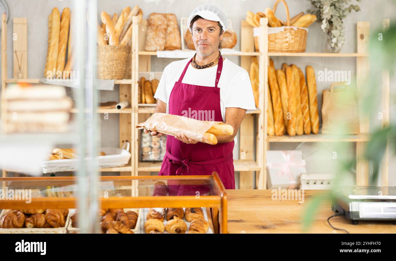 Middle-aged salesman holding paper bag with baguettes in bakery Stock ...