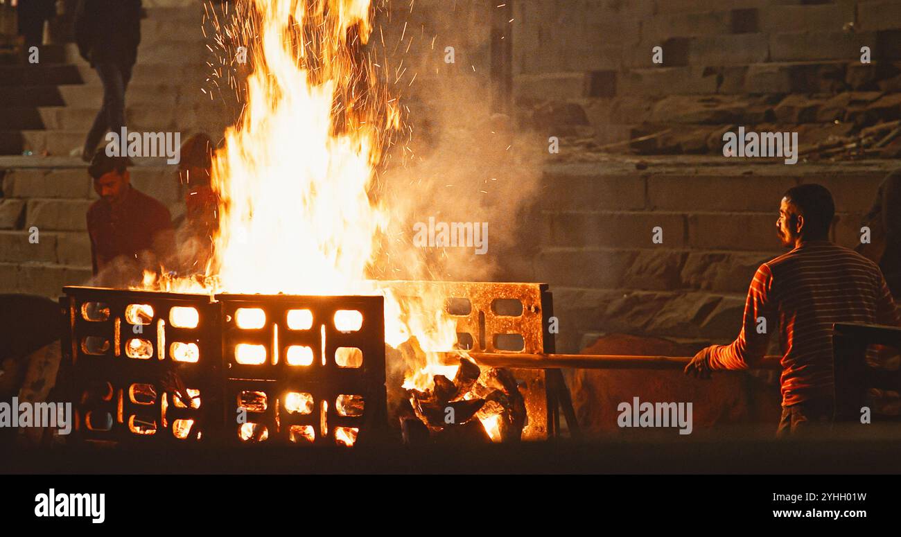 Varanasi, Uttar Pradesh, India. Indian worker man helps a fire burn ...