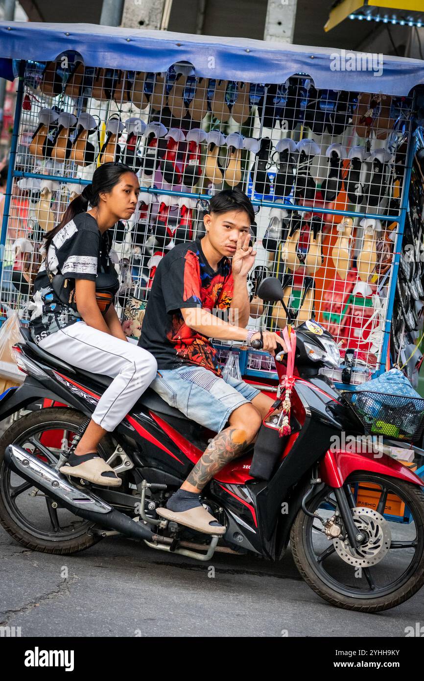 A beautiful young Thai girl rides on the back of a motorbike behind her ...