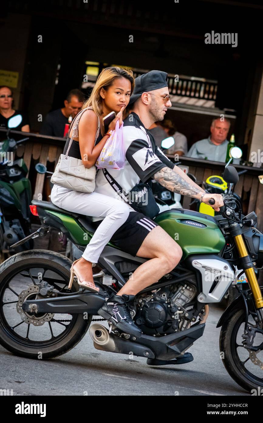 A beautiful young Thai girl rides on the back of a motorbike behind her ...