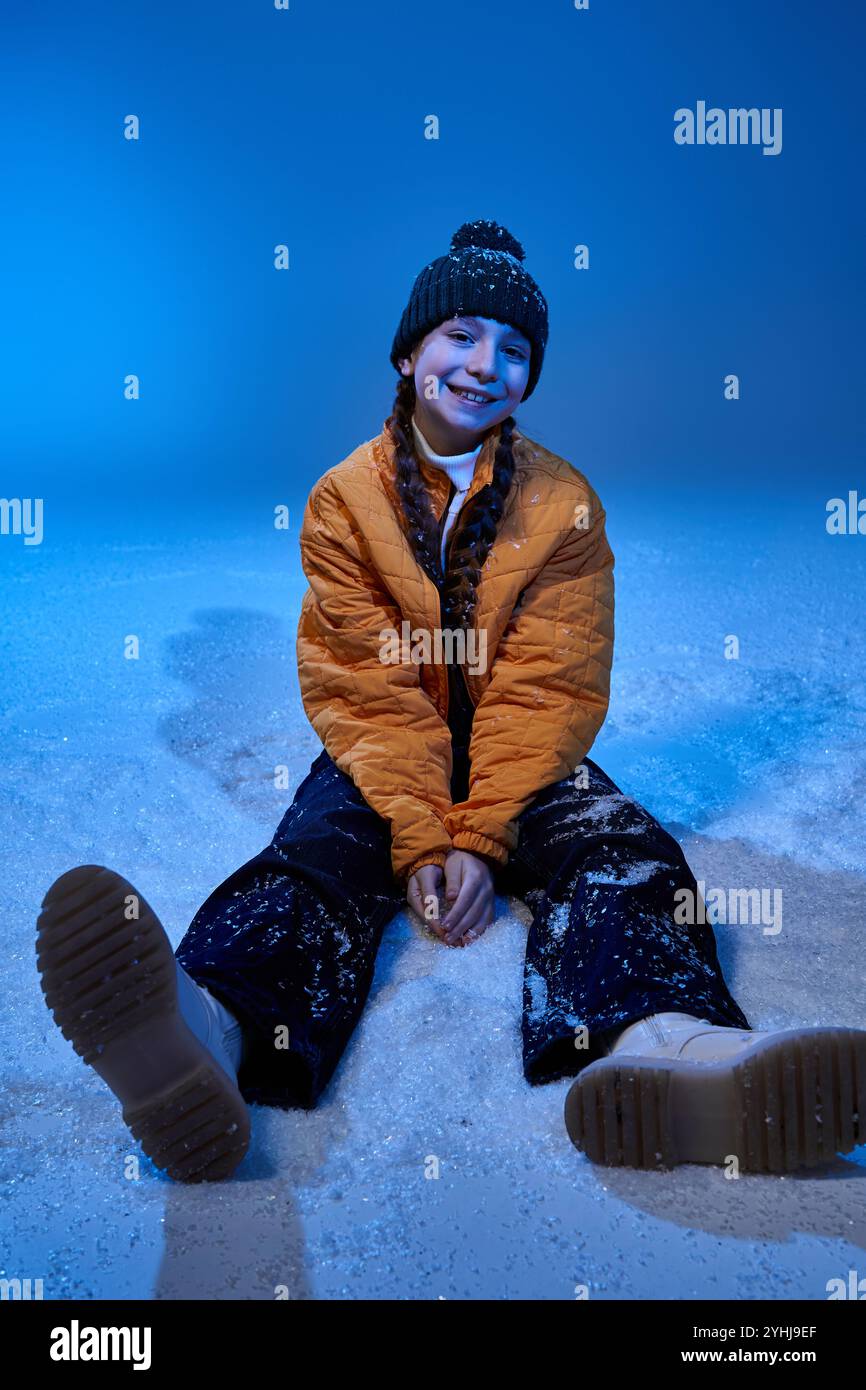 A happy girl in cozy winter clothing sits on the snow, smiling brightly ...