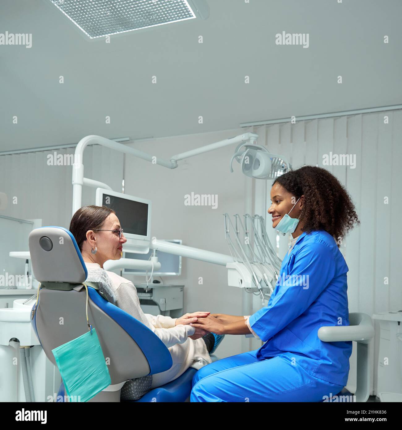 Dentist sits beside patient in dental chair, gently holding her hand ...