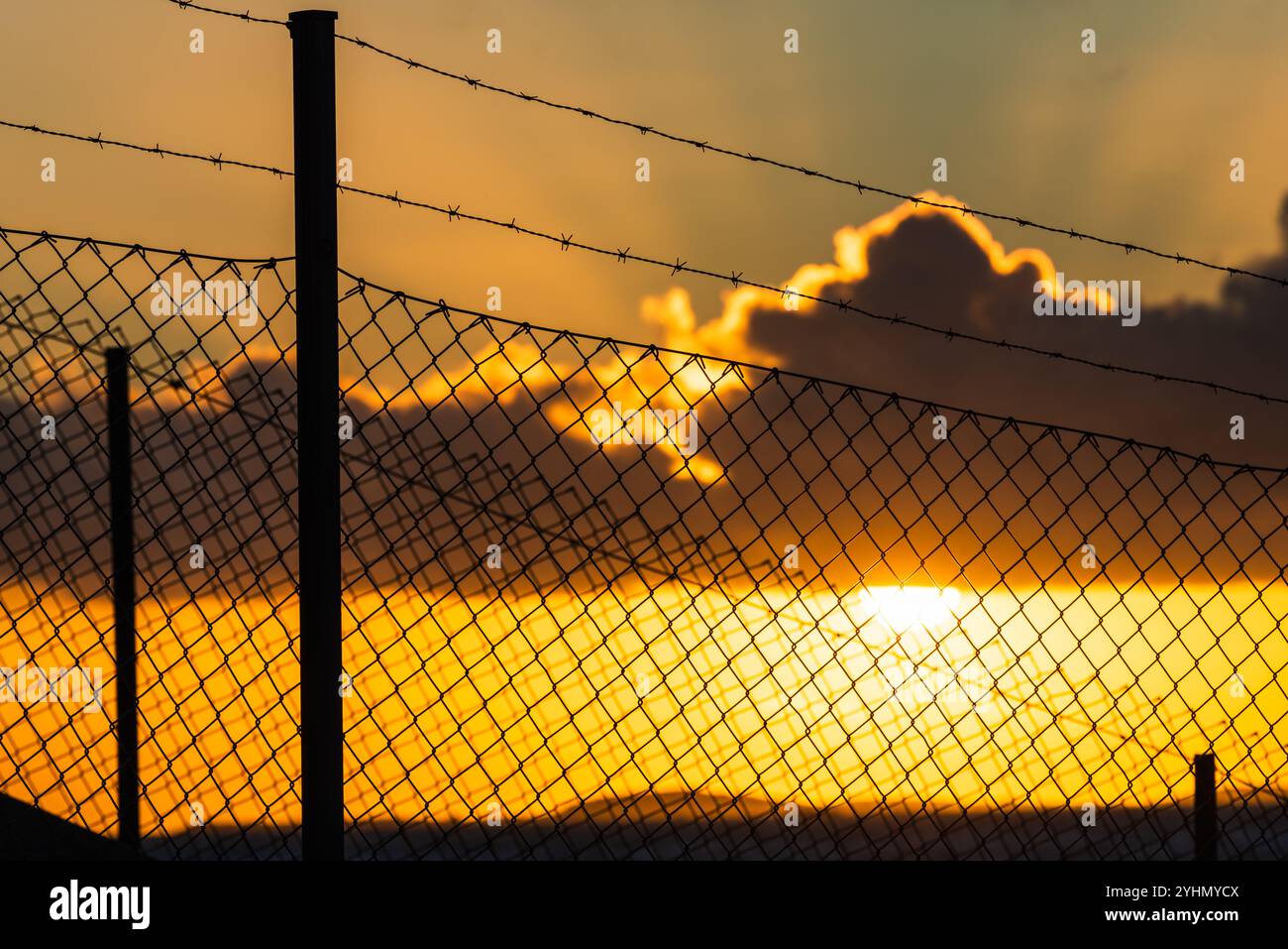 The sun sets behind a barbed wire fence, casting vibrant orange and ...