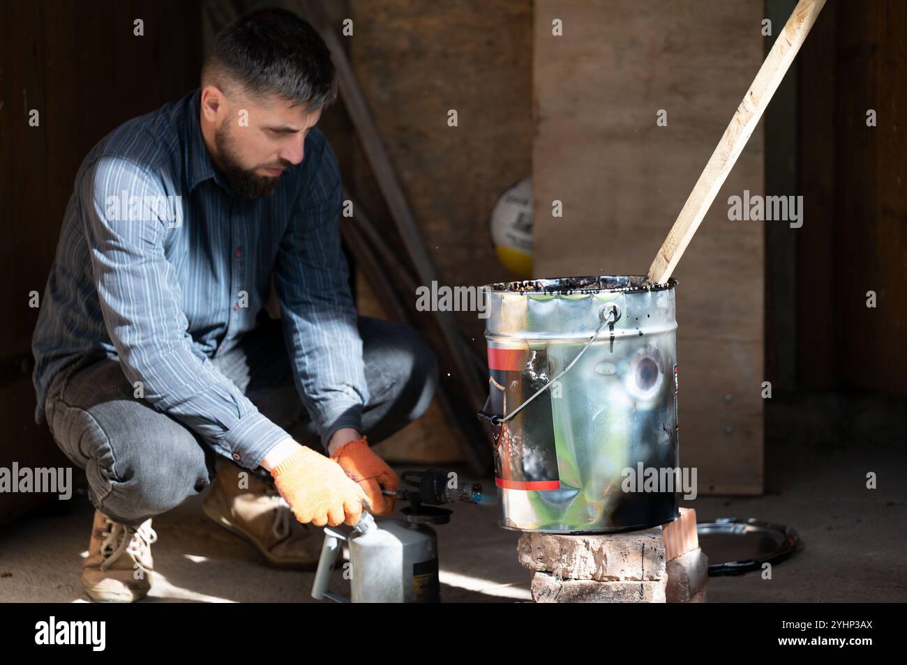 A man stirs liquid bitumen in a bucket with a wooden stick ...
