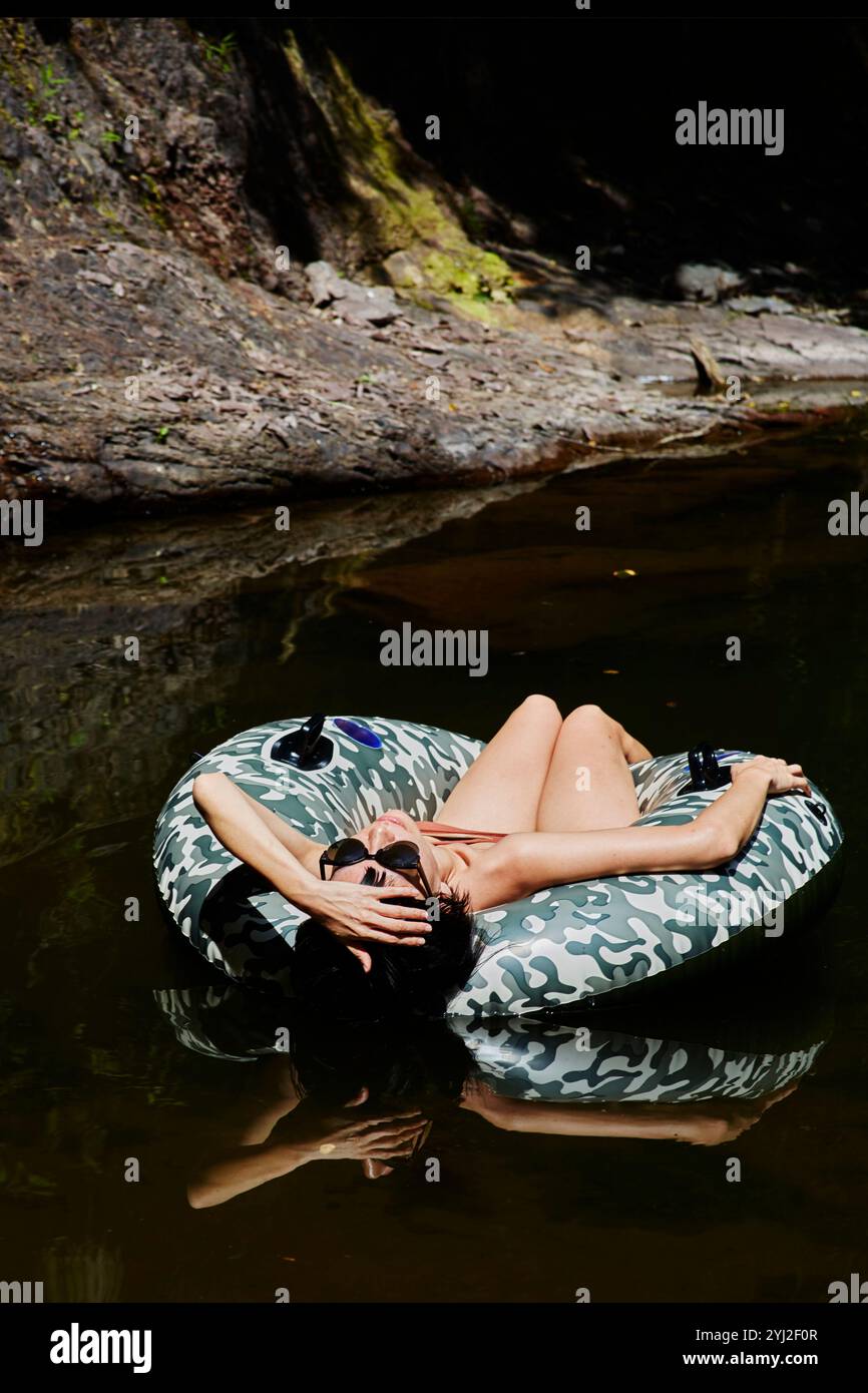 Woman relaxing on an inflatable ring in a serene forest river, basking in sunlight with her ...