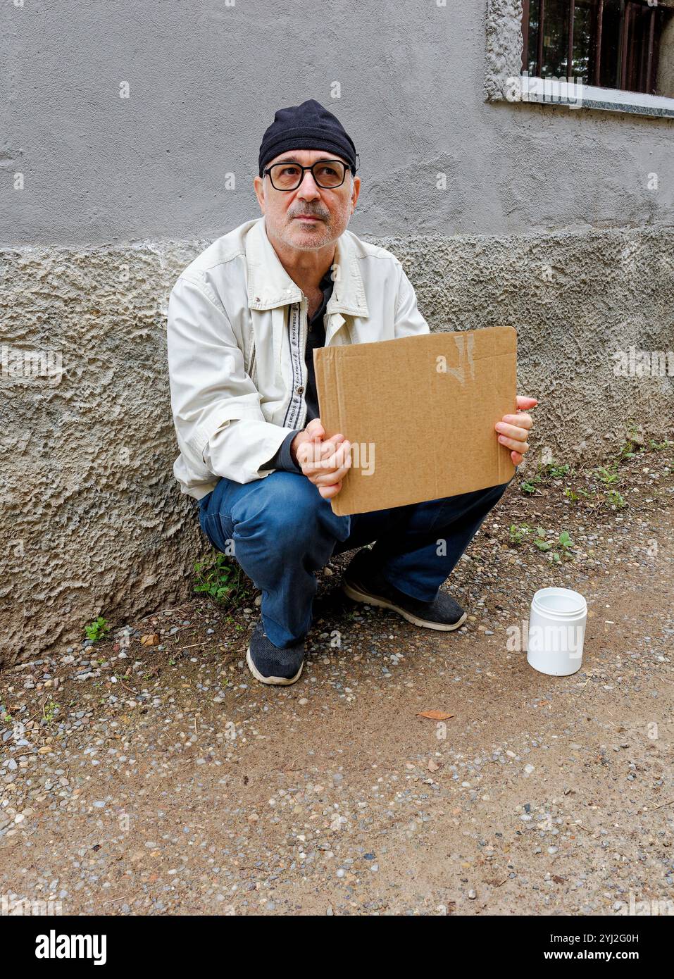 Middle-aged man squatting on the sidewalk holding a cardboard sign with ...