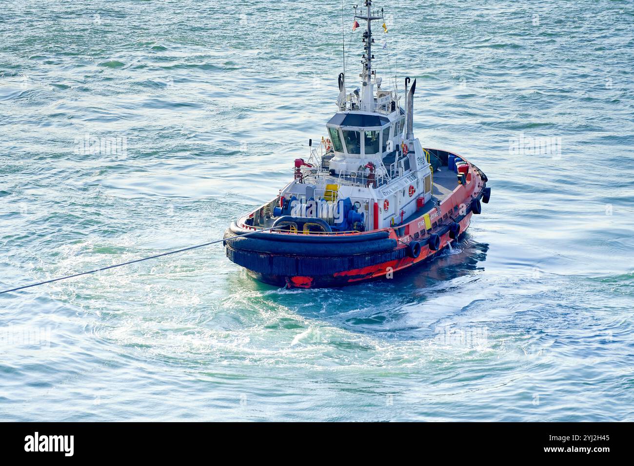 Red and blue tugboat towing a large unseen vessel on the open sea ...