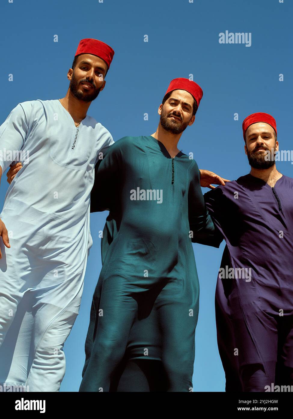 Three men in traditional attire and red fez hats posing against a clear ...