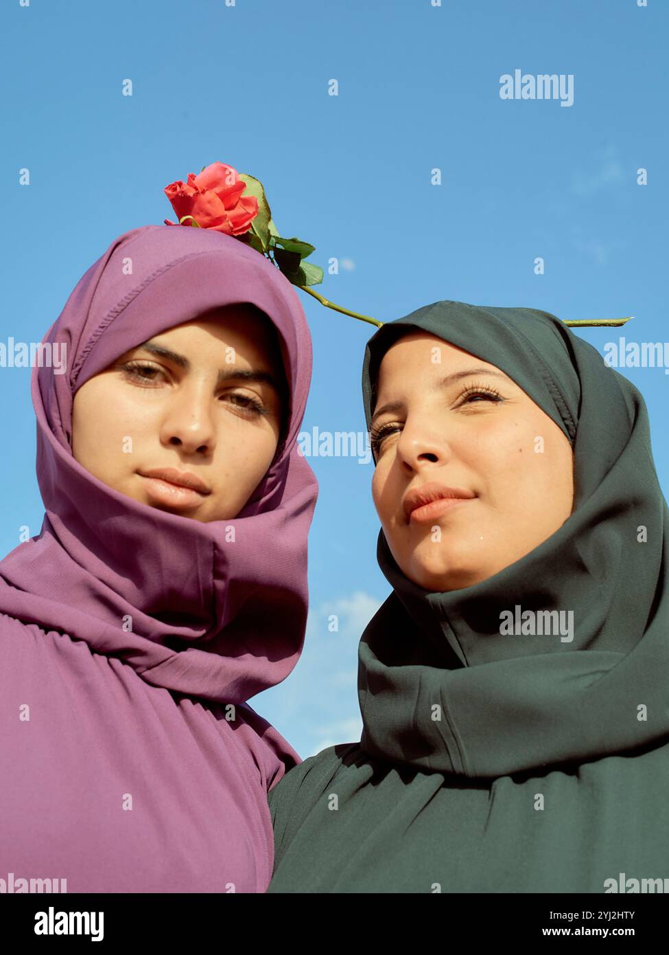 Two women wearing colorful hijabs pose with a rose against a clear blue ...