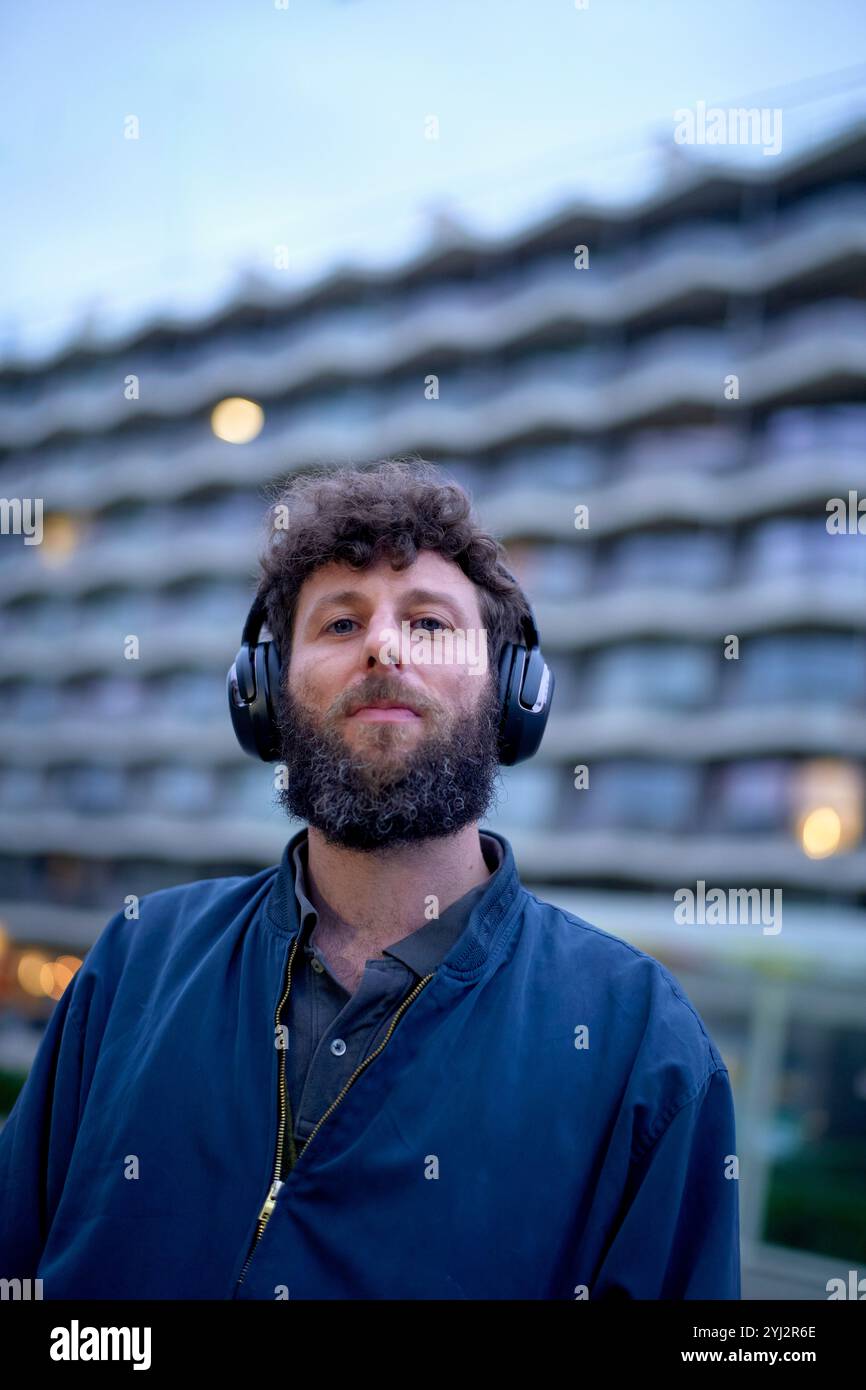 Bearded man with headphones standing against a blurred building ...