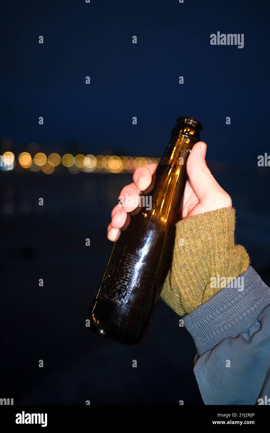 Hand holding a brown beer bottle against a blurred nighttime beach ...