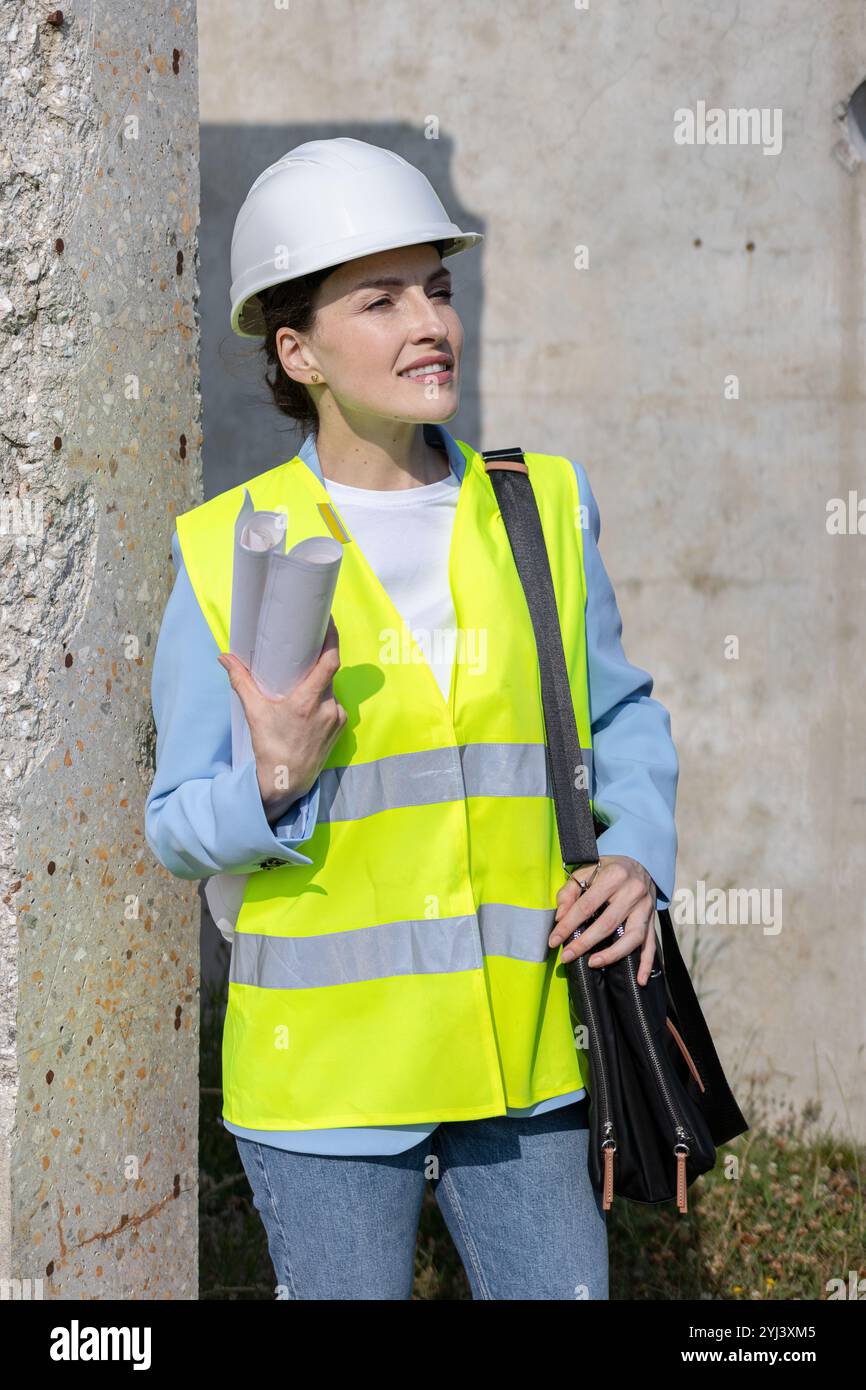 Confident architect wearing safety helmet and reflective vest holding blueprints while ...