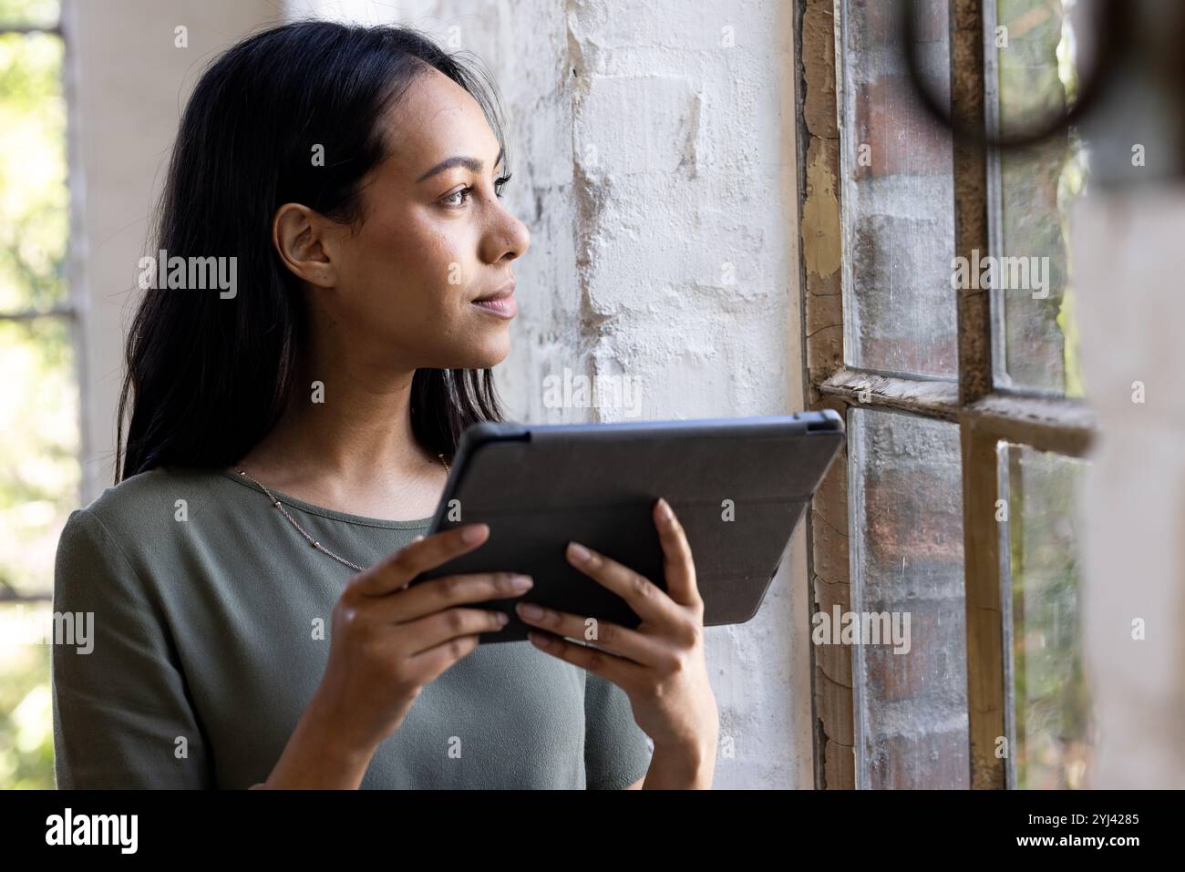 Holding tablet, woman gazing thoughtfully out window in home setting ...