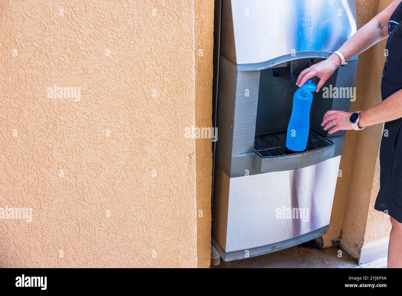 Woman collecting ice cubes from dispenser to cool water bottle for cold ...