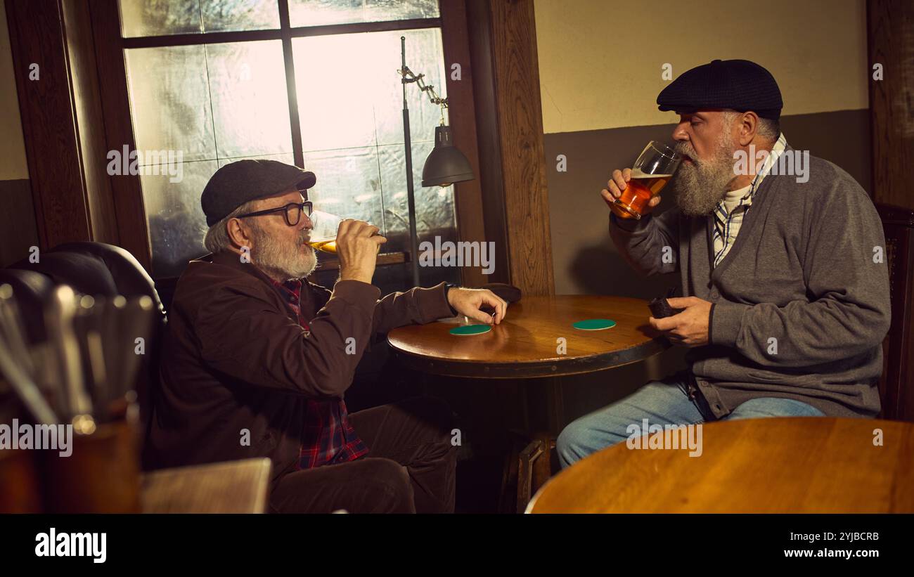 Two elderly men drinking beer at wooden table in rustic atmospheric bar ...