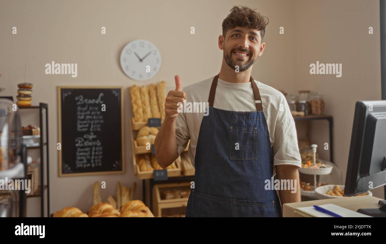 Young man in bakery shop interior with apron giving thumbs-up next to ...