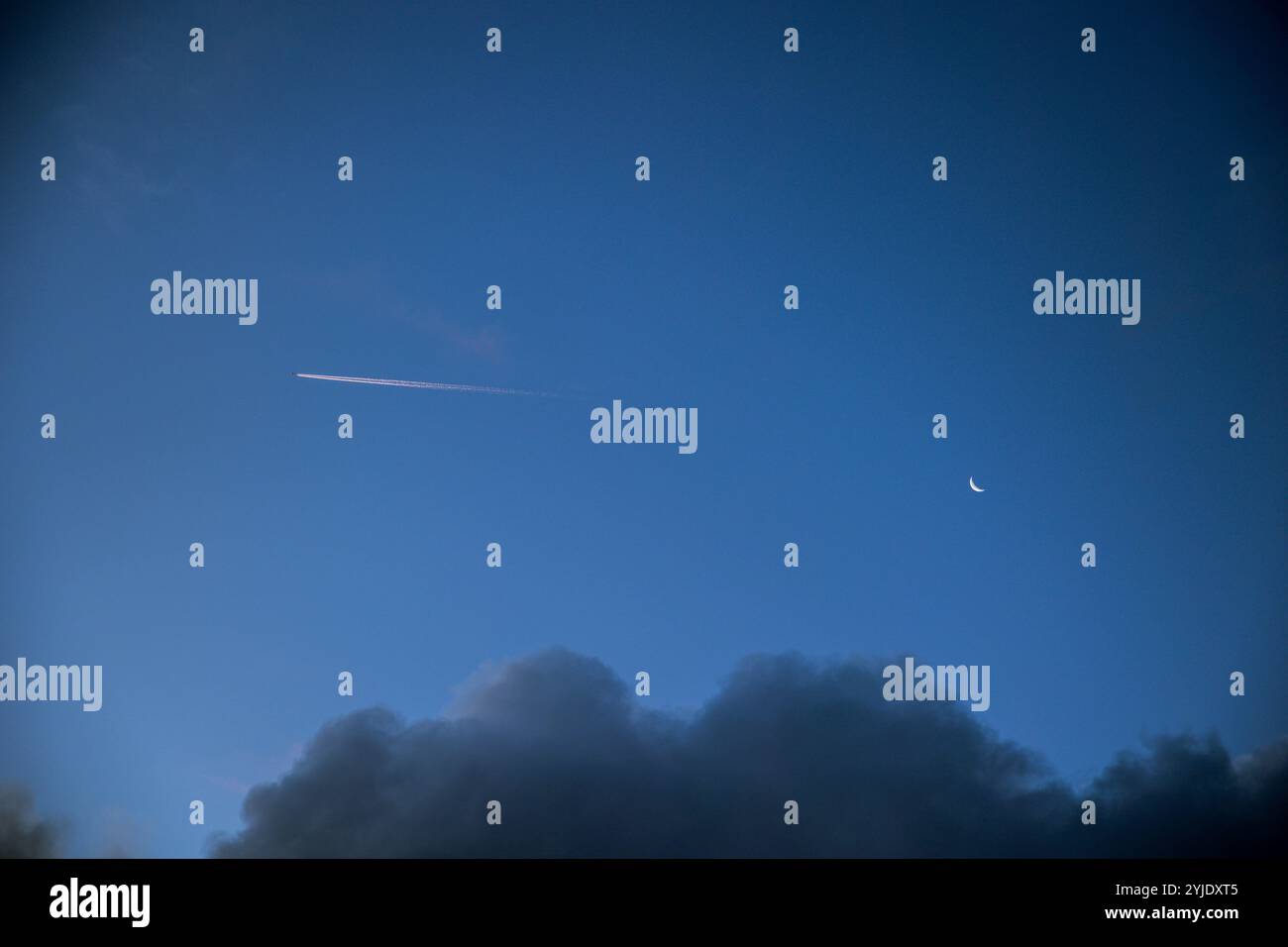Blue sky with clouds and waning moon with a plane with a white wake ...