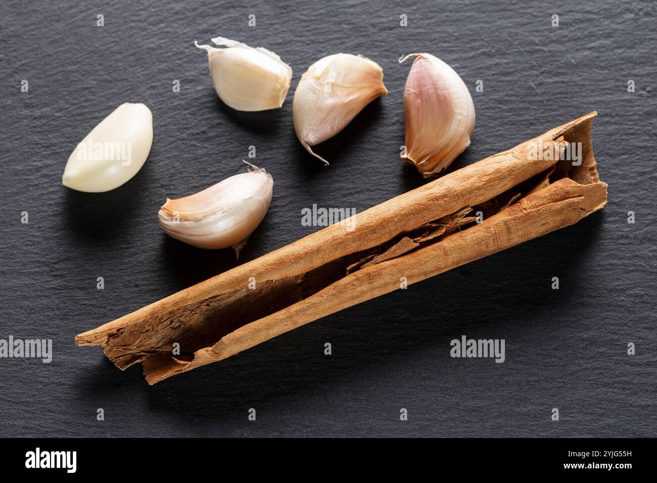 Four garlic cloves and a cinnamon stick lie on a black slate surface ...