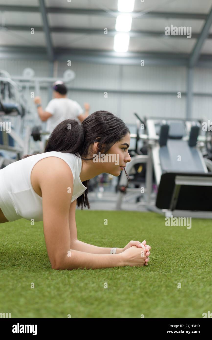 A young woman performs a plank exercise on a green turf floor in a gym ...