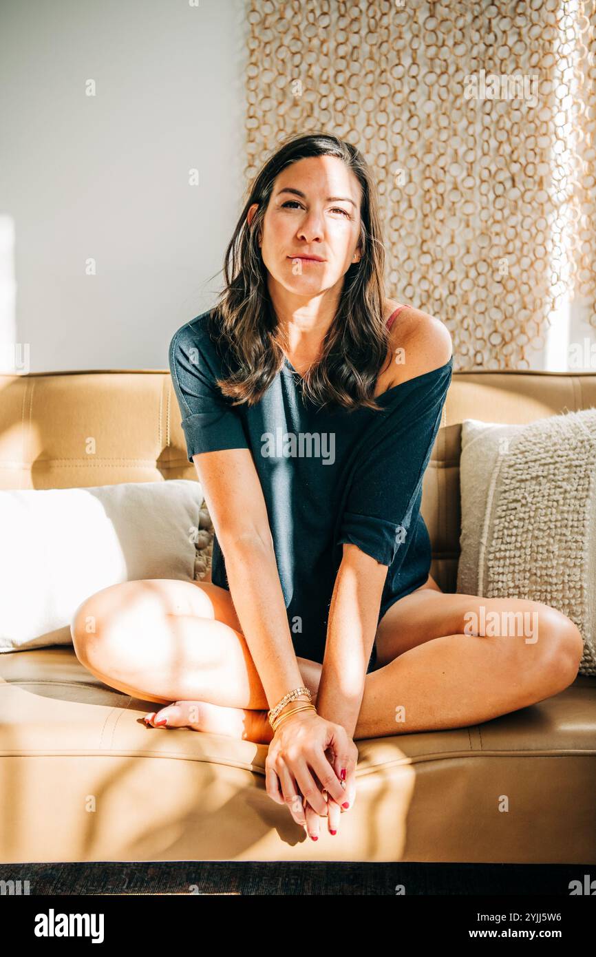 Woman sitting cross-legged on couch, gazing thoughtfully in sunlight ...