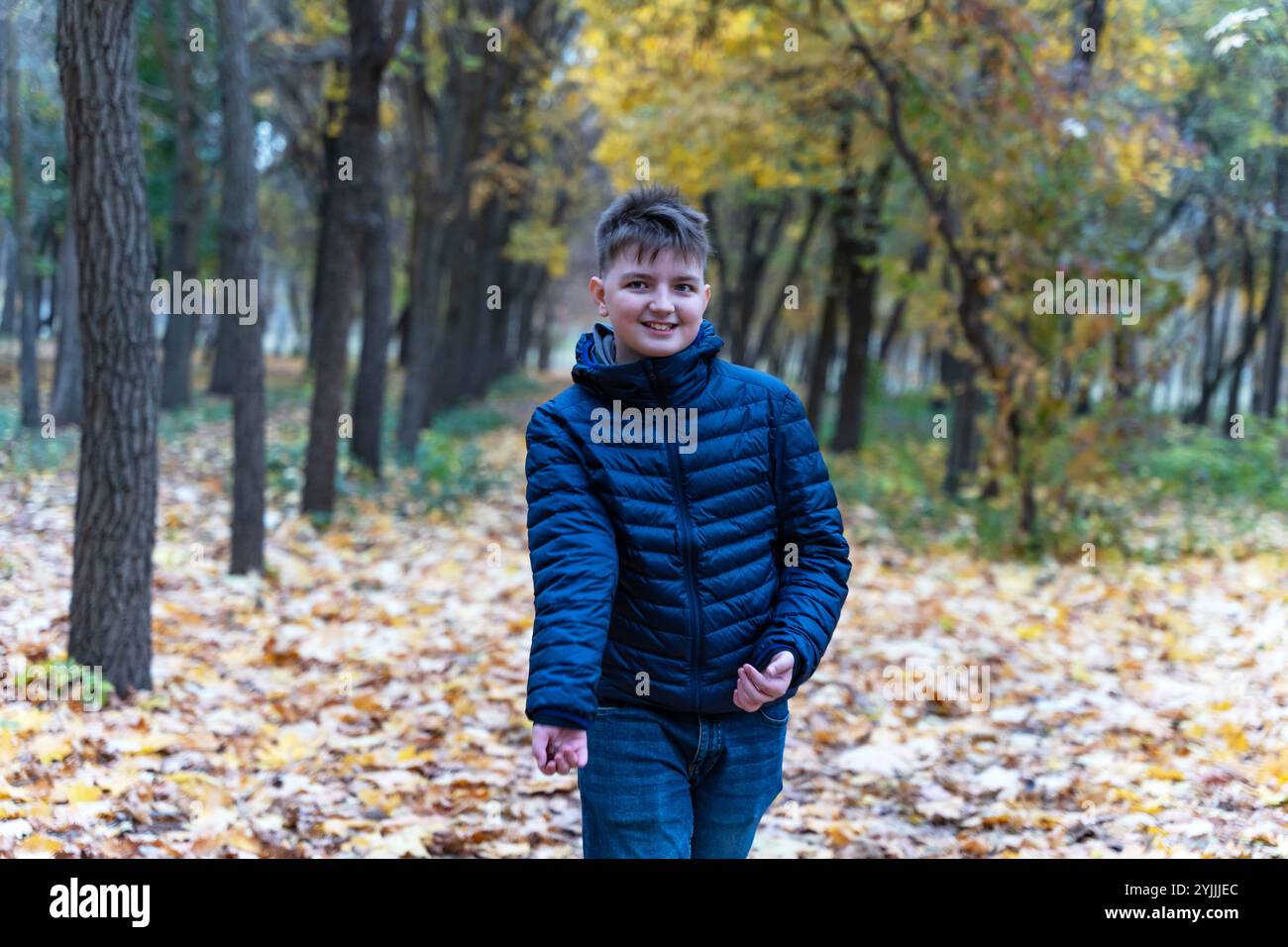 In an autumn park, a 12-year-old smiling white boy in a blue jacket and ...